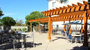 Área de pátio externa com mesas e cadeiras, estrutura de pérgola e guarda-sóis para sombra.