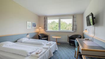 Quarto de hotel com duas camas, uma escrivaninha, uma cadeira, uma mesinha, uma televisão e uma janela com cortinas.