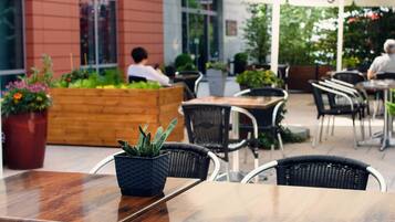 Um café ao ar livre com mesas de madeira e cadeiras de vime preto, um vaso com plantas verdes e um edifício com grandes janelas ao fundo.