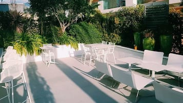 Um pátio externo com cadeiras e mesas brancas, rodeado por vegetação e um edifício com varandas.