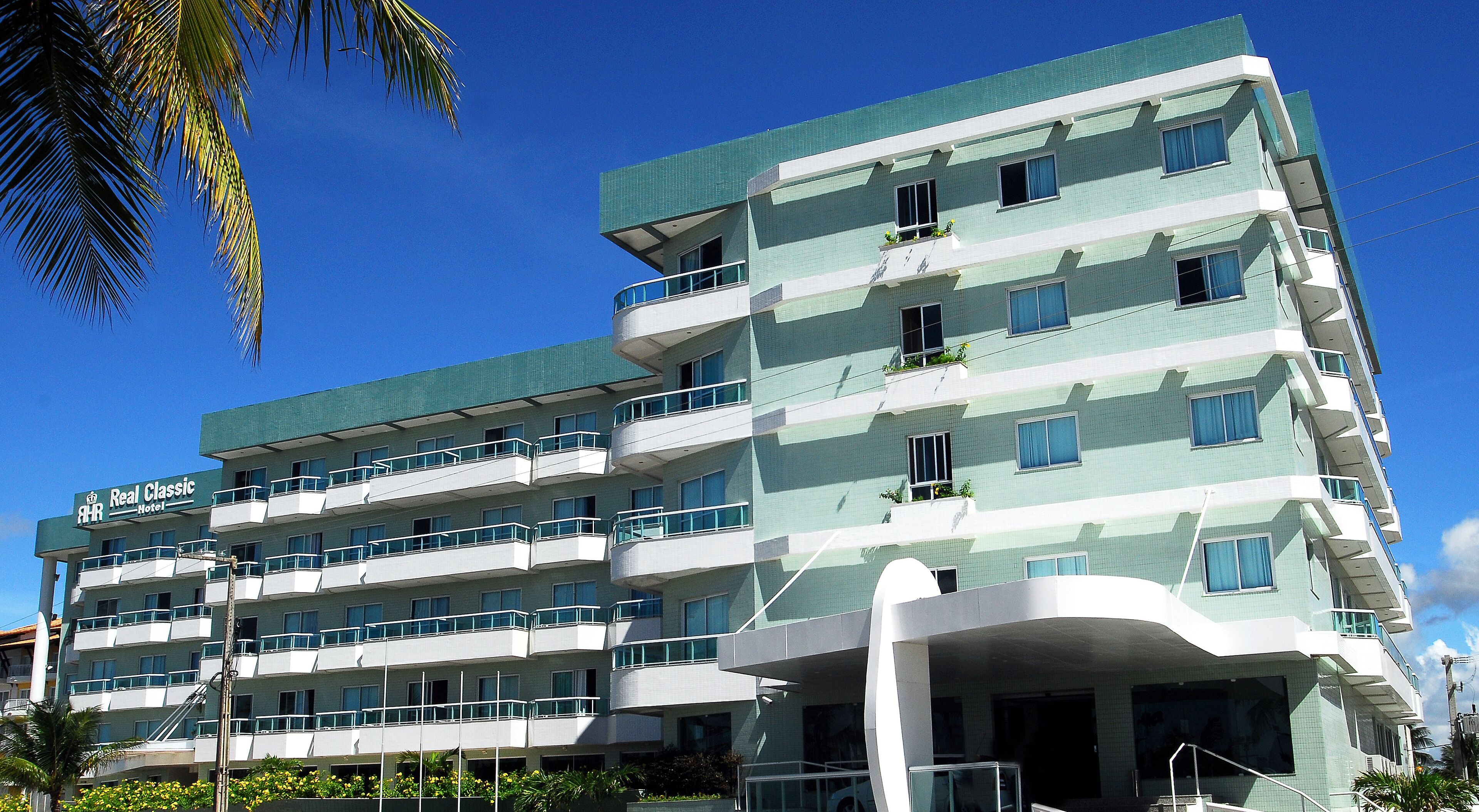 Um hotel de vários andares com varandas, uma placa indicando ser o Real Classic Hotel, e um céu azul claro ao fundo.
