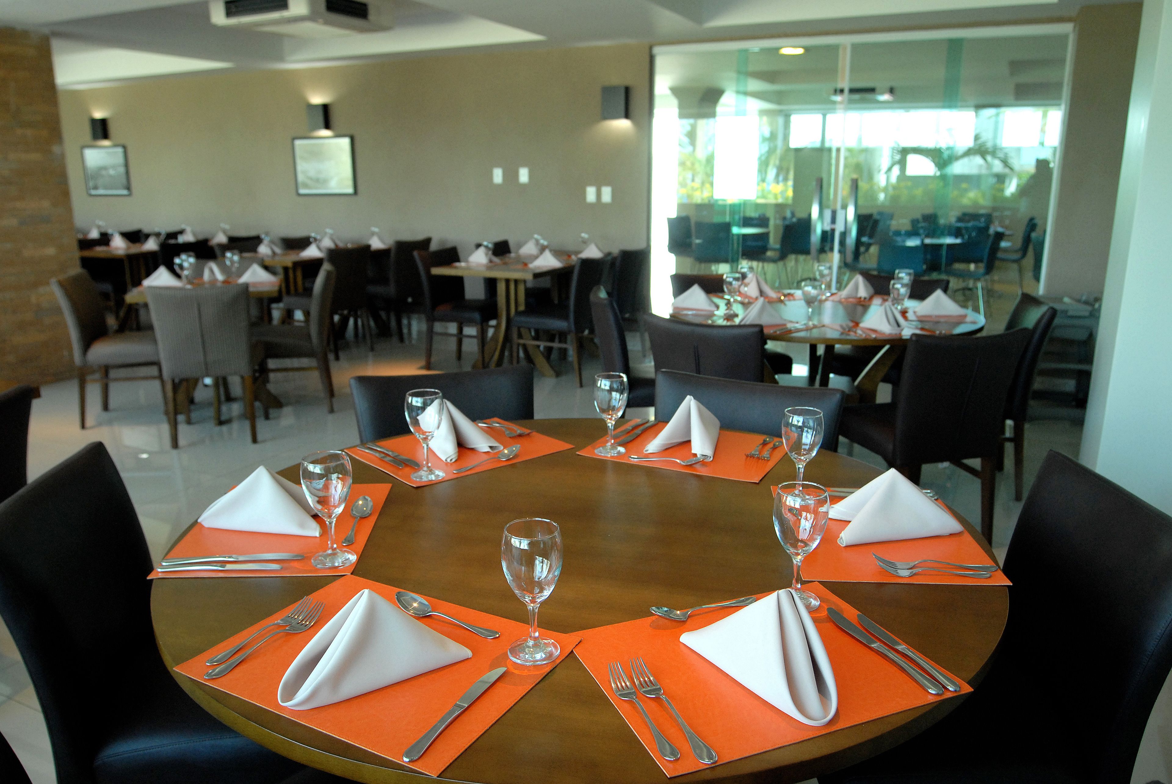 Restaurante com mesas redondas preparadas para refeições, com jogos americanos laranja e talheres organizados.