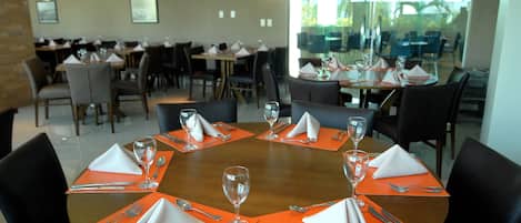 Restaurante com mesas redondas preparadas para refeições, com jogos americanos laranja e talheres organizados.