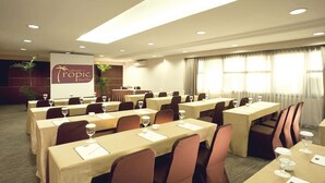 Uma sala de conferência com mesas preparadas para uma reunião, uma tela de projetor e um banner com a inscrição "Grand Tropic Suites Hotel".