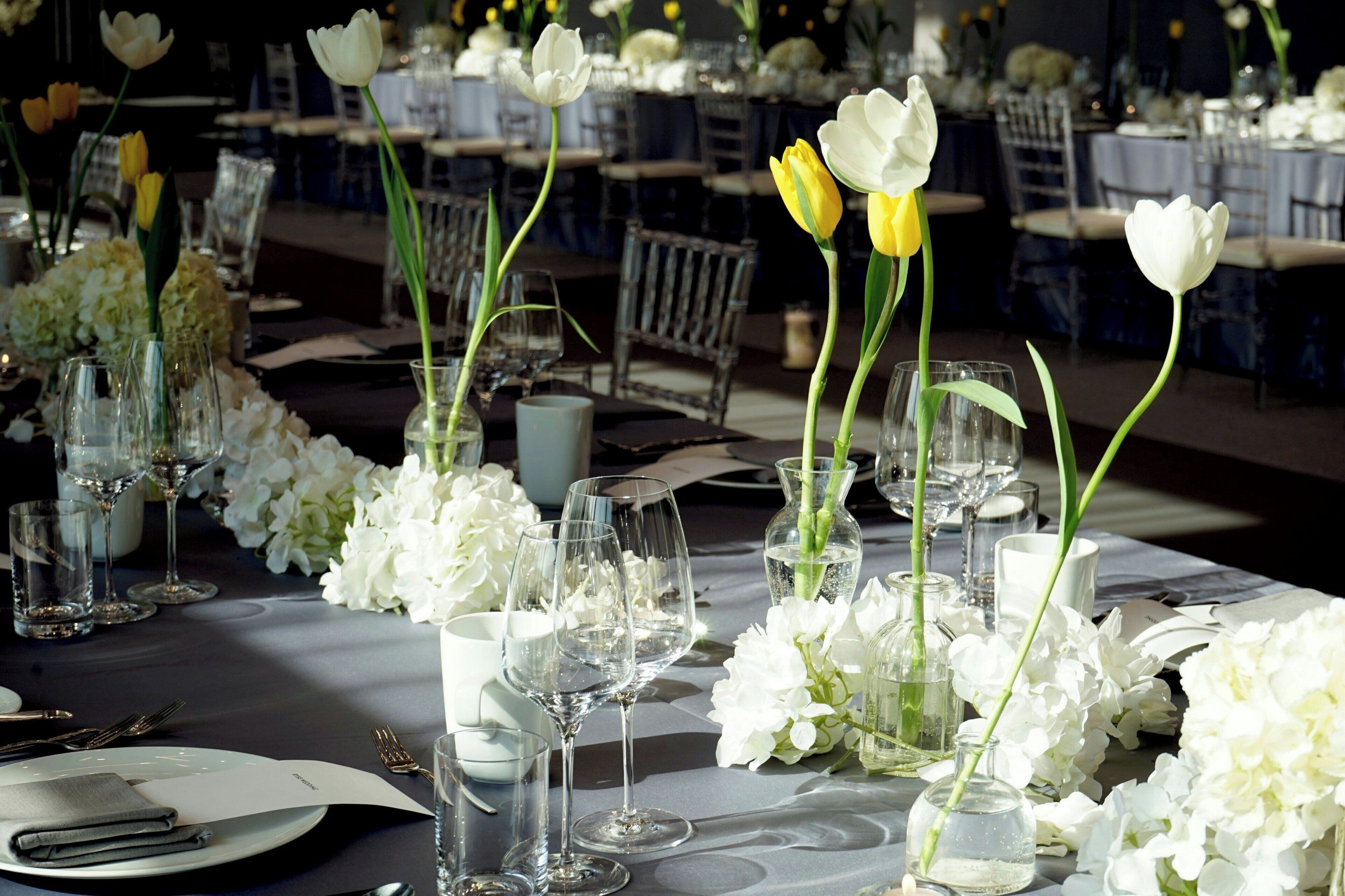 Salão de banquetes com mesas postas para refeição, com tulipas brancas e amarelas e flores brancas em vasos de vidro.