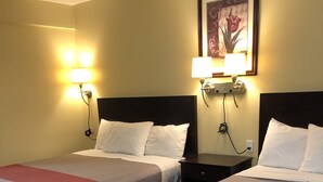 Quarto de hotel com duas camas, cada uma com manta vermelha e branca, uma mesinha de cabeceira e duas luminárias fixadas na parede.