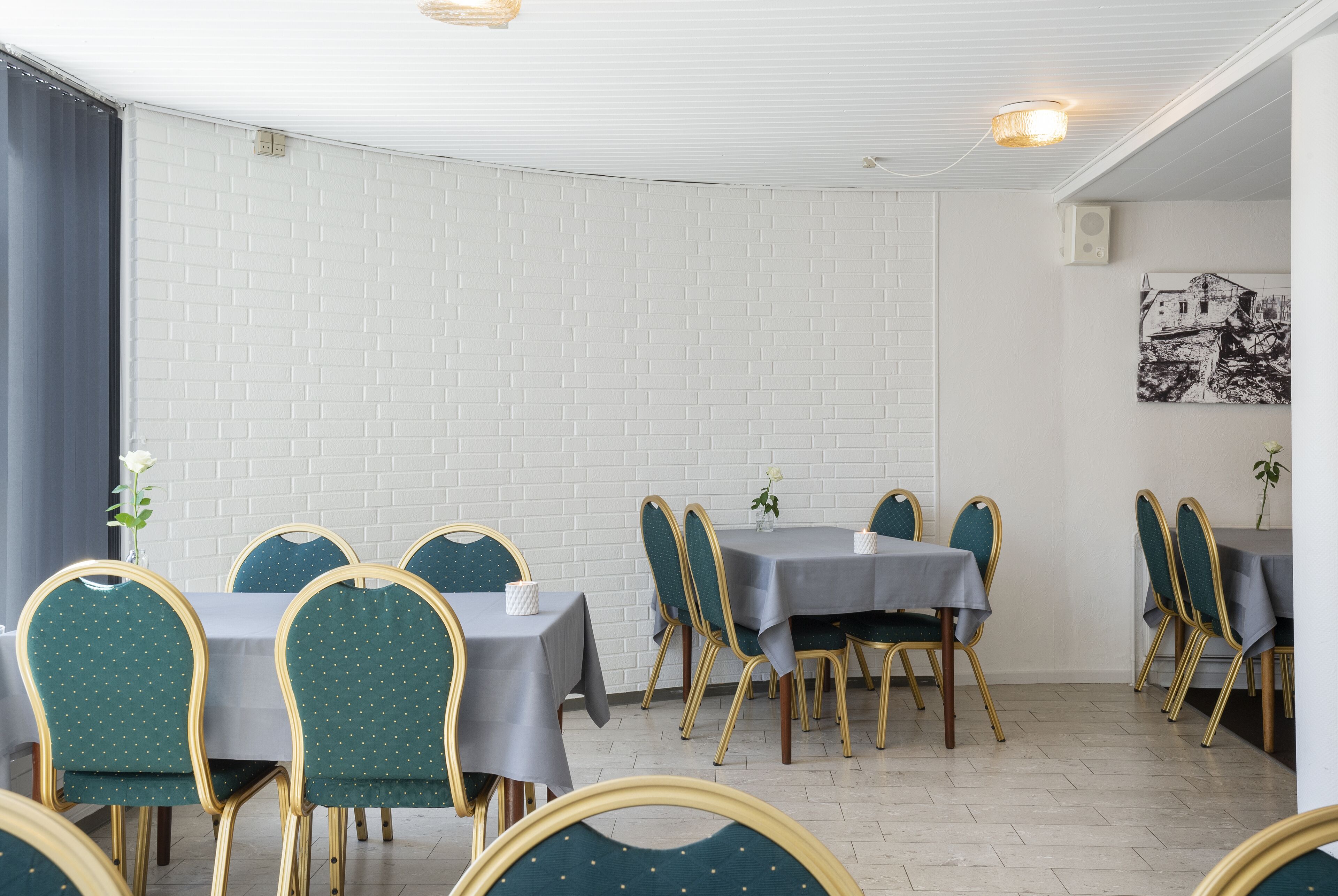 Uma área de refeições com paredes de tijolo branco, cadeiras estofadas verdes com detalhes dourados e toalhas de mesa cinzentas.