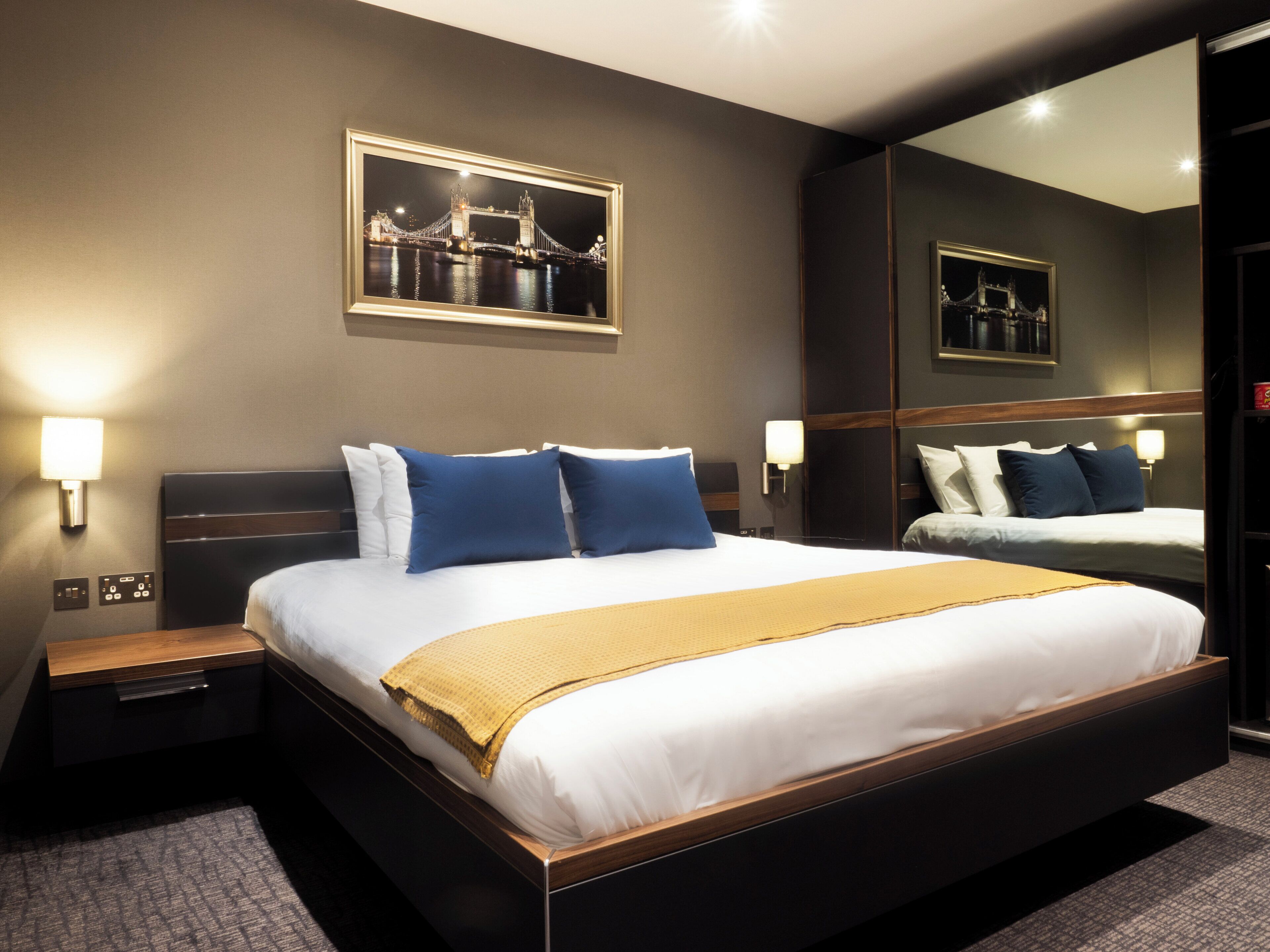 Quarto de hotel com uma cama grande, duas mesas de cabeceira, um criado-mudo, uma luminária fixada na parede e um quadro com a imagem de uma ponte.