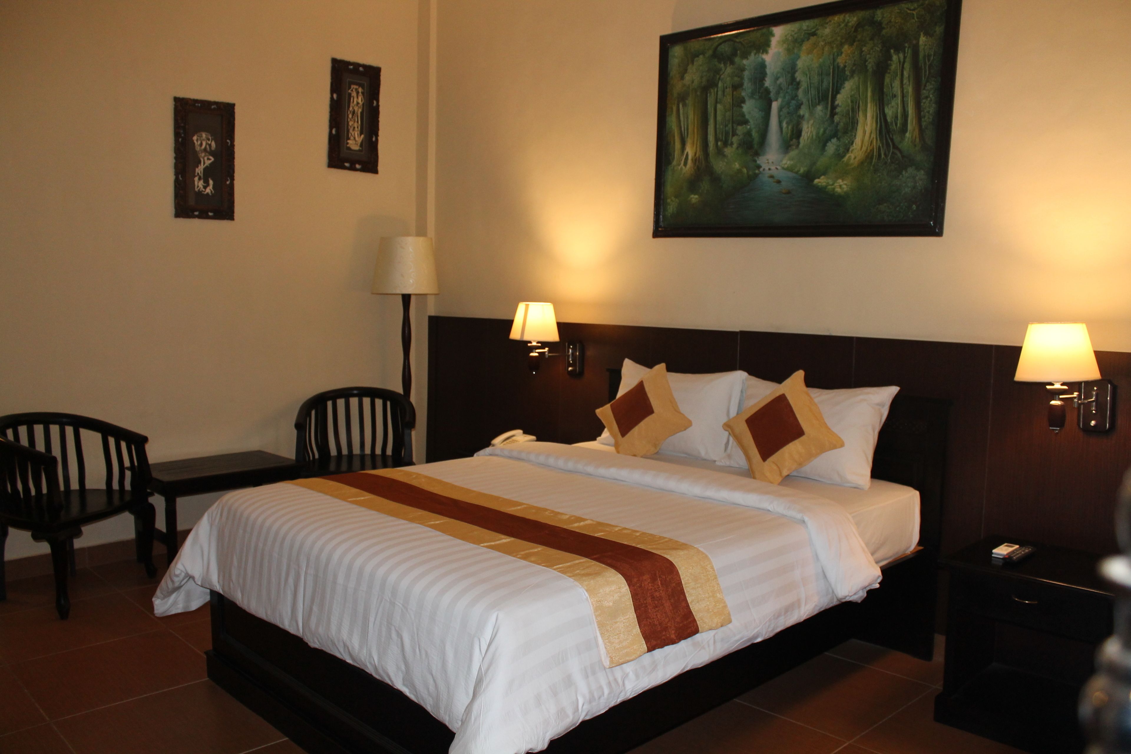 Uma cama bem arrumada com edredom branco e marrom, duas luminárias de cabeceira, um encosto, um criado-mudo e duas cadeiras.