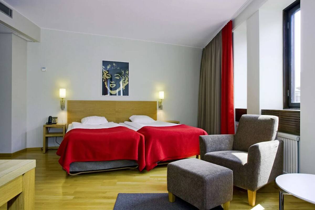Quarto de hotel com uma cama, duas mesas de cabeceira, uma cadeira, uma mesinha e uma janela com cortinas.