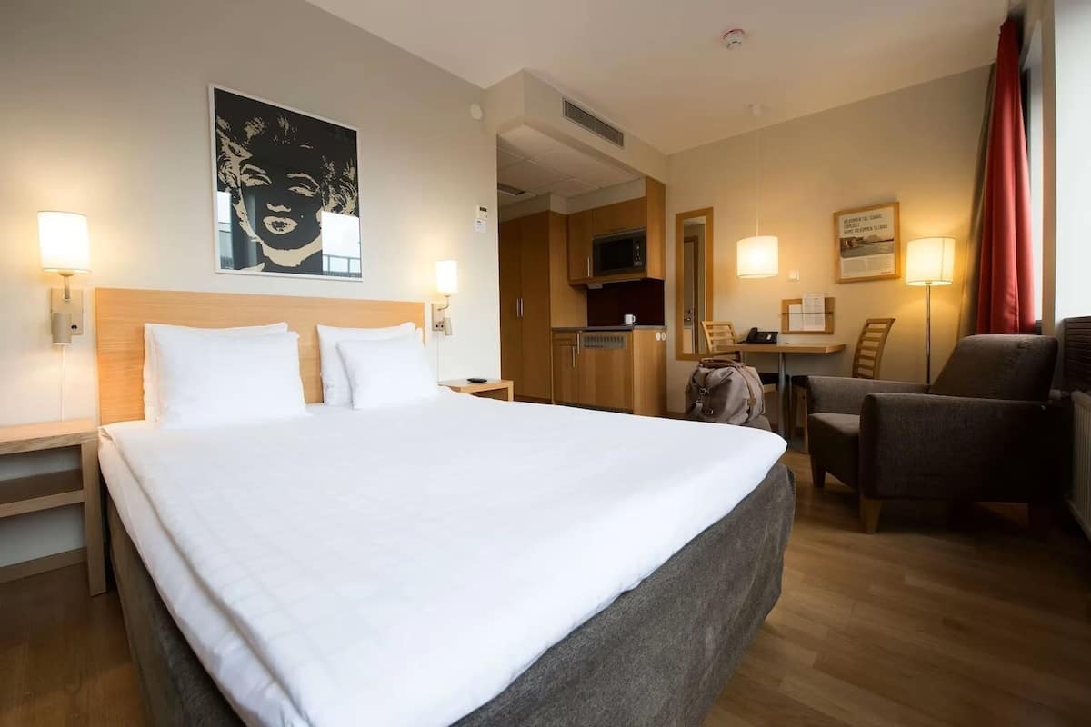 Quarto de hotel com uma cama grande, uma escrivaninha, uma cadeira, micro-ondas e uma pequena cozinha equipada.