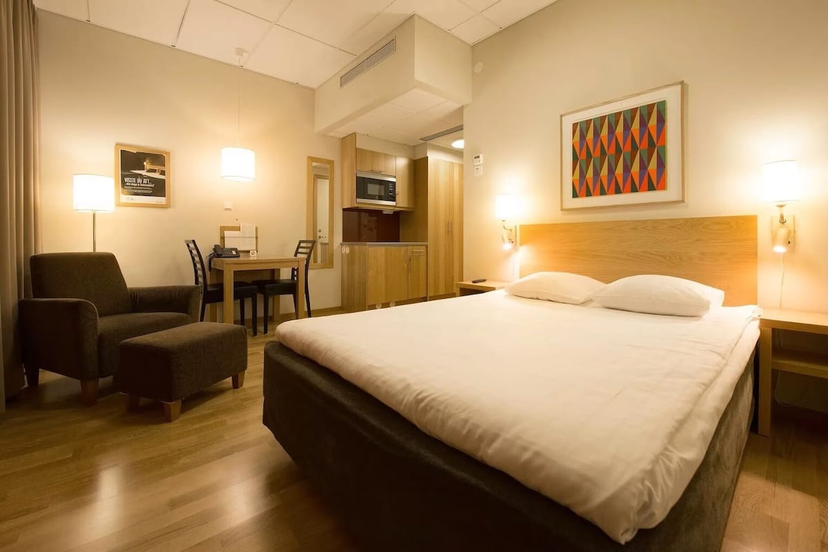 Quarto de hotel com cama, escrivaninha, cadeira, micro-ondas e um quadro na parede.