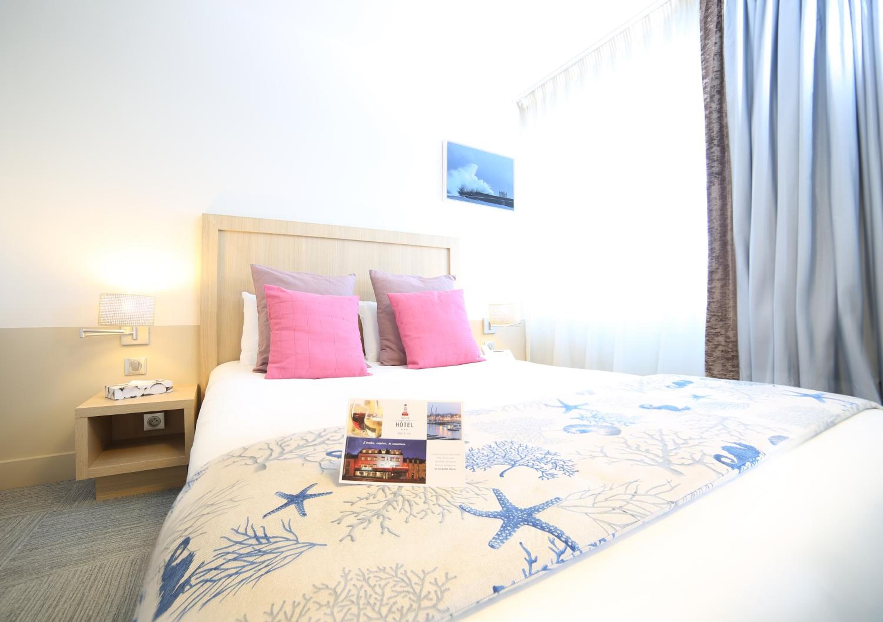 Quarto de hotel com uma cama, dois travesseiros cor-de-rosa, uma mesa de cabeceira com abajur, um quadro na parede e uma janela com cortinas.