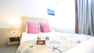 Quarto de hotel com uma cama, dois travesseiros cor-de-rosa, uma mesa de cabeceira com abajur, um quadro na parede e uma janela com cortinas.