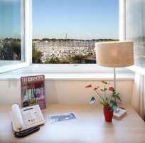 Quarto de hotel com vista para um porto, uma mesa com telefone, uma revista e um vaso de plantas.