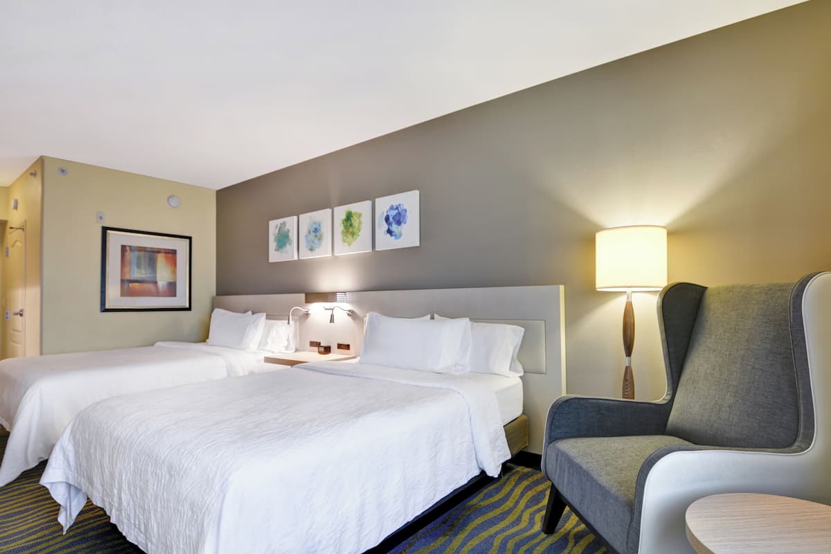 Quarto de hotel com duas camas, uma cadeira, uma luminária e uma obra de arte na parede.