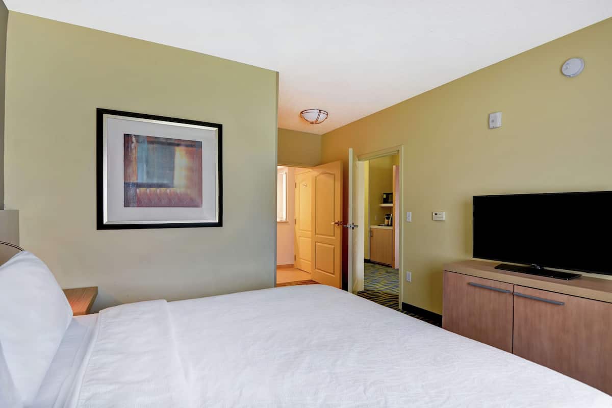 Quarto de hotel com uma cama, uma televisão sobre um armário de madeira, um quadro pendurado na parede e uma porta que dá para um corredor.