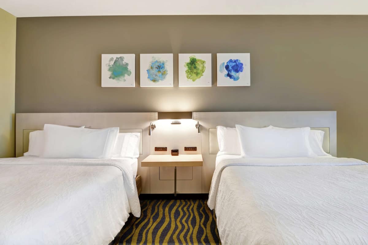 Quarto de hotel com duas camas, uma mesinha e quatro obras de arte abstratas nas paredes.