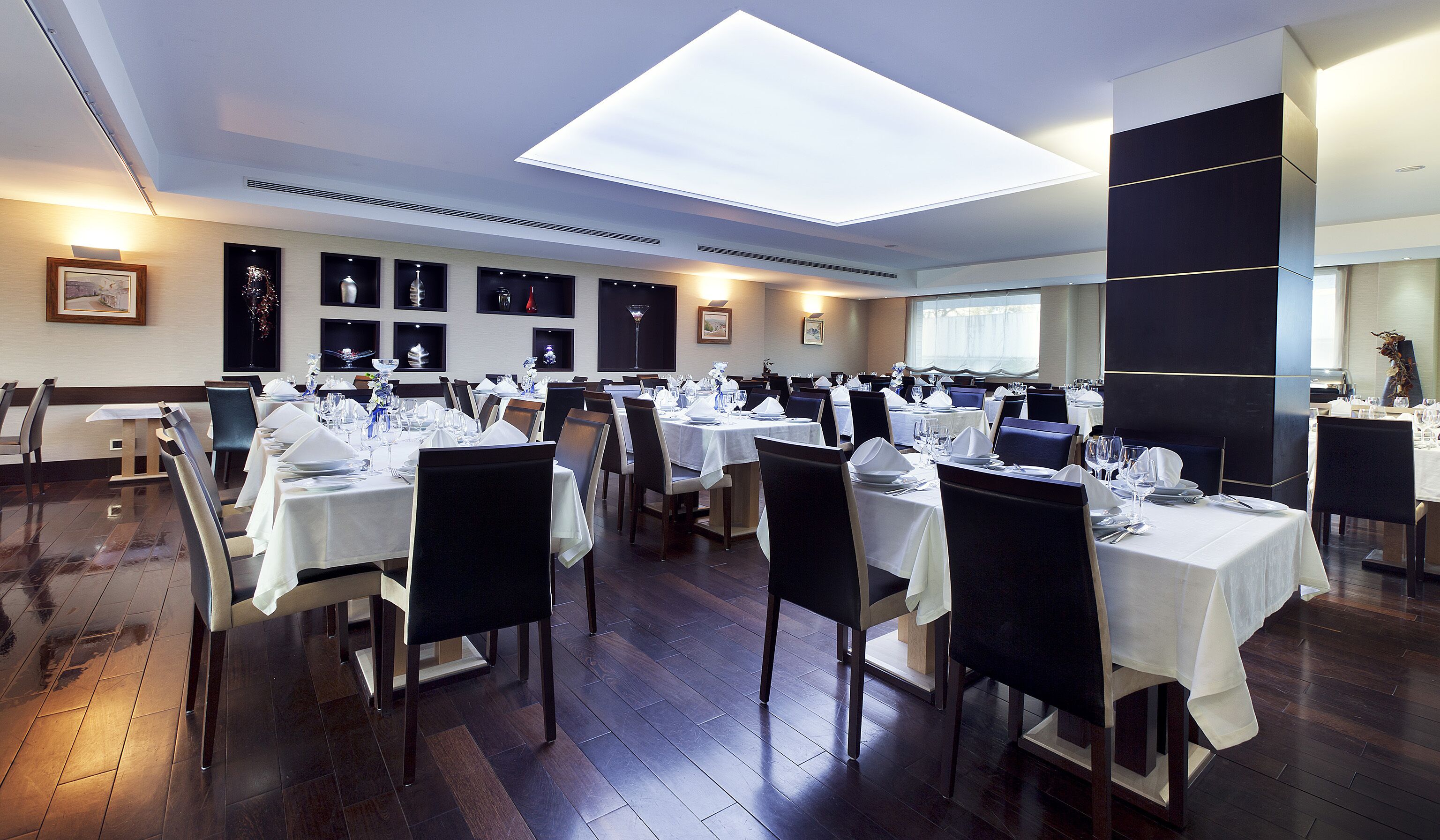 Uma ampla sala de jantar com várias mesas montadas para um evento formal, com teto alto e iluminação moderna.