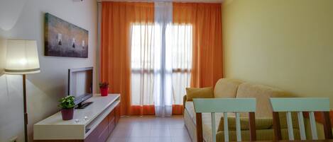 Uma sala de estar com televisão, luminária, sofá, mesa de jantar com frutas e janela com cortinas laranja.