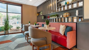 Uma área de estar moderna com um sofá vermelho, duas mesas redondas e cadeiras. Há prateleiras com objetos decorativos e plantas.