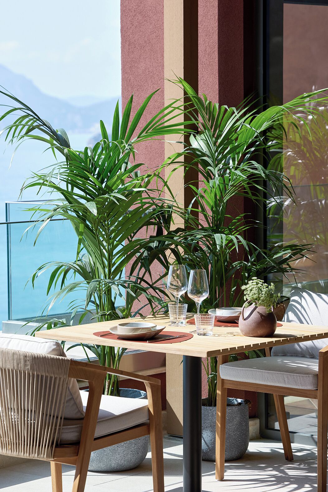 Uma mesa de madeira posta para refeição, com plantas em vasos, um copo de água e uma taça de vinho.