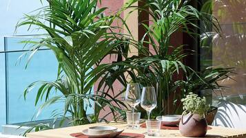 Uma mesa de madeira posta para refeição, com plantas em vasos, um copo de água e uma taça de vinho.