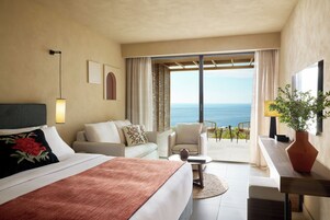 Quarto de hotel com cama, sofá, poltrona, mesinha e vista para o mar por grandes janelas.