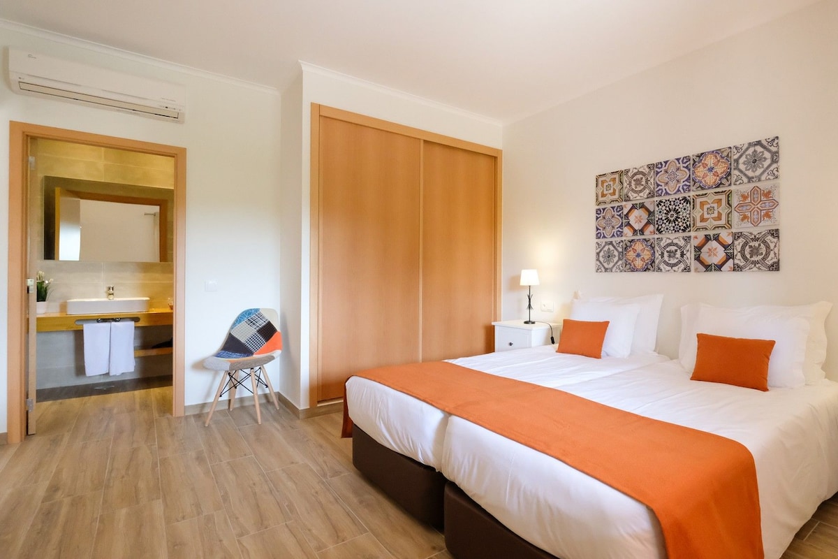 Quarto de hotel com cama, mesa de cabeceira, cadeira, armário e banheiro visível por uma porta aberta.