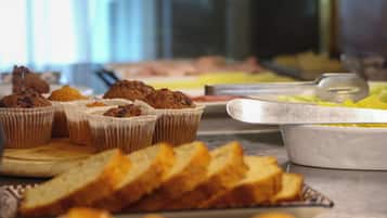 Mesa de buffet com diversos alimentos, incluindo muffins, pão e uma tigela branca.