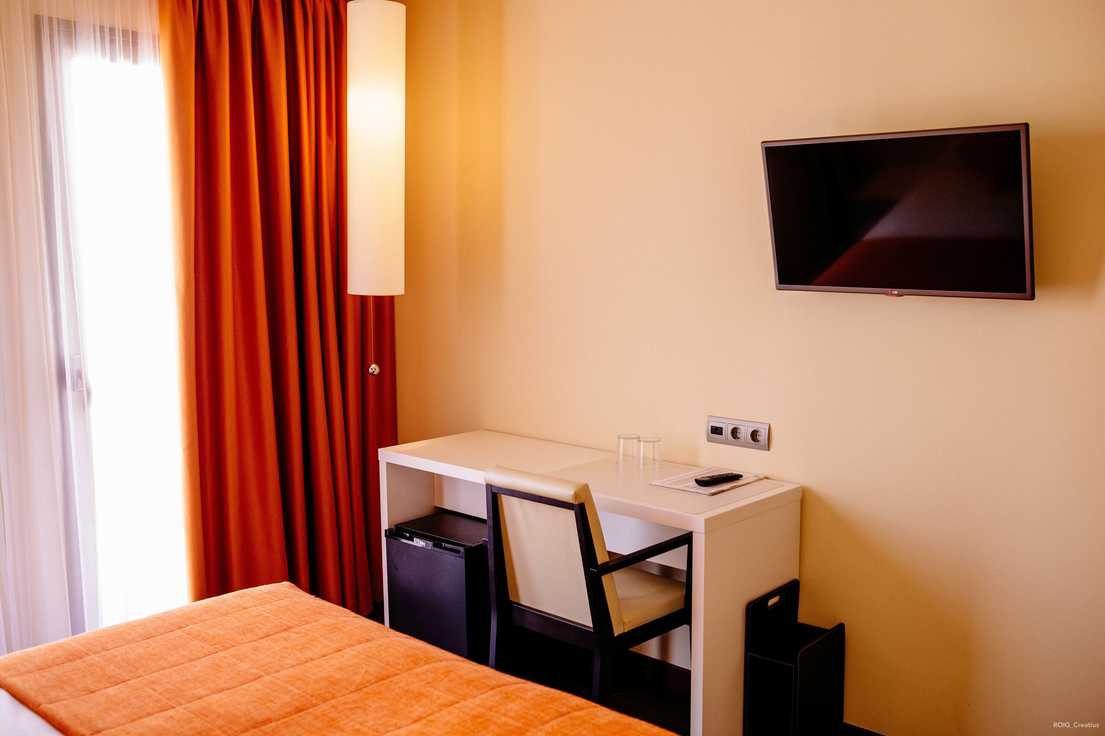 Quarto de hotel com cama, escrivaninha com cadeira, televisão e uma pequena lixeira.