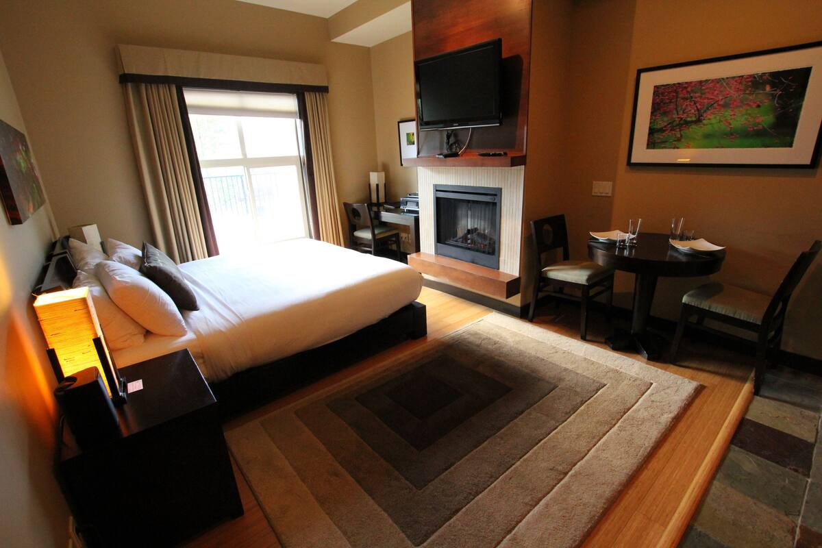 Quarto de hotel com uma cama grande, televisão de tela plana, lareira e área de jantar com mesa e cadeiras.