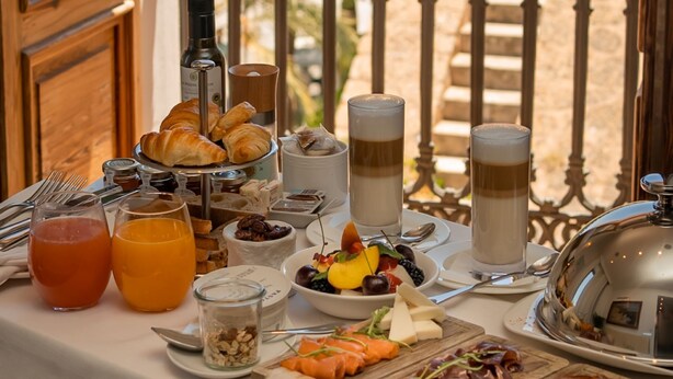 Mesa de café da manhã com croissants, frutas e bebidas, com vista para o mar e palmeiras.