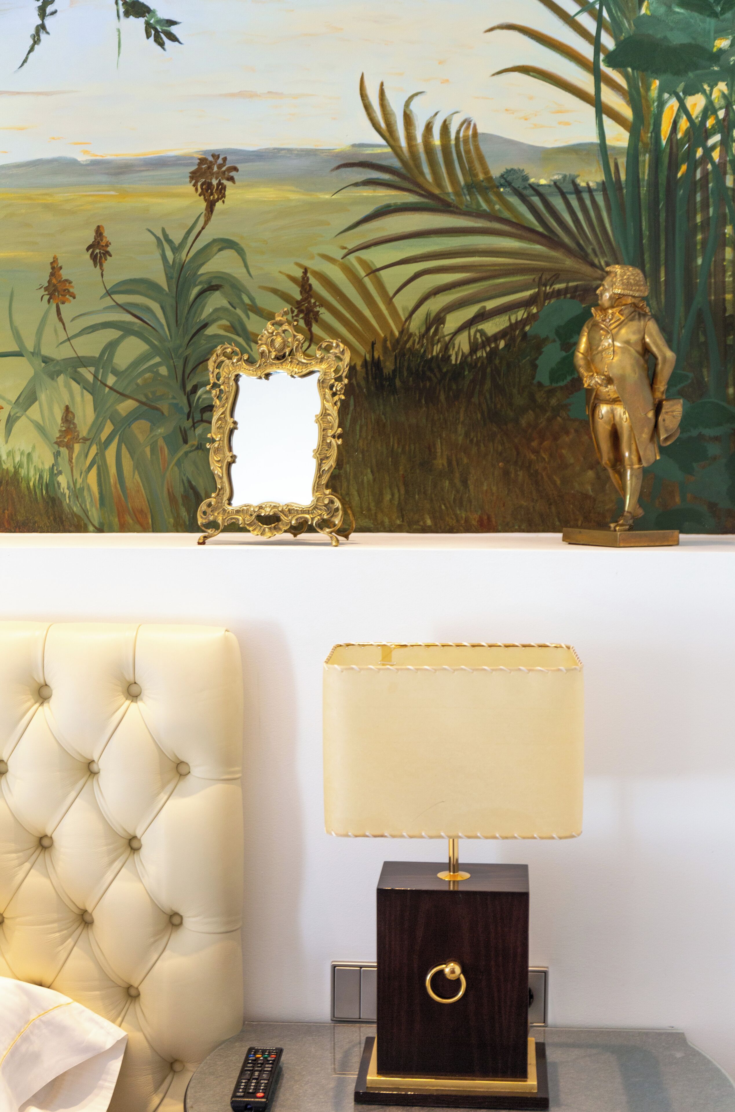 Quarto com cabeceira estofada, mesa de cabeceira com abajur, espelho emoldurado e uma estátua.