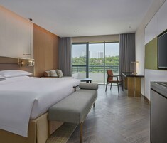 Quarto de hotel com uma cama grande, uma escrivaninha, uma cadeira e um banco. Há uma janela com vista para edifícios e vegetação.