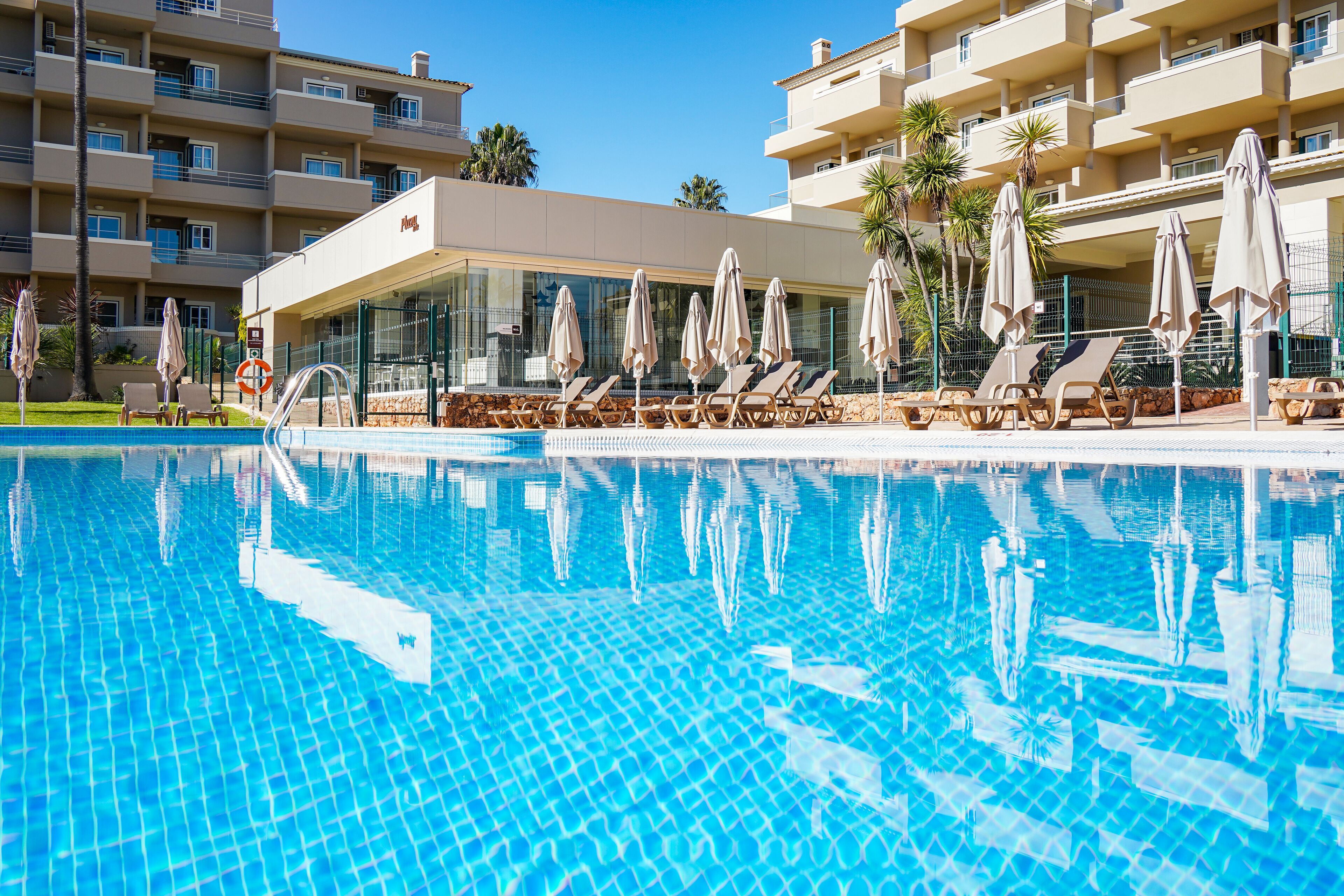 Piscina com espreguiçadeiras e guarda-sóis, cercada por um edifício com varandas e um céu claro.