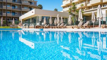 Piscina com espreguiçadeiras e guarda-sóis, cercada por um edifício com varandas e um céu claro.