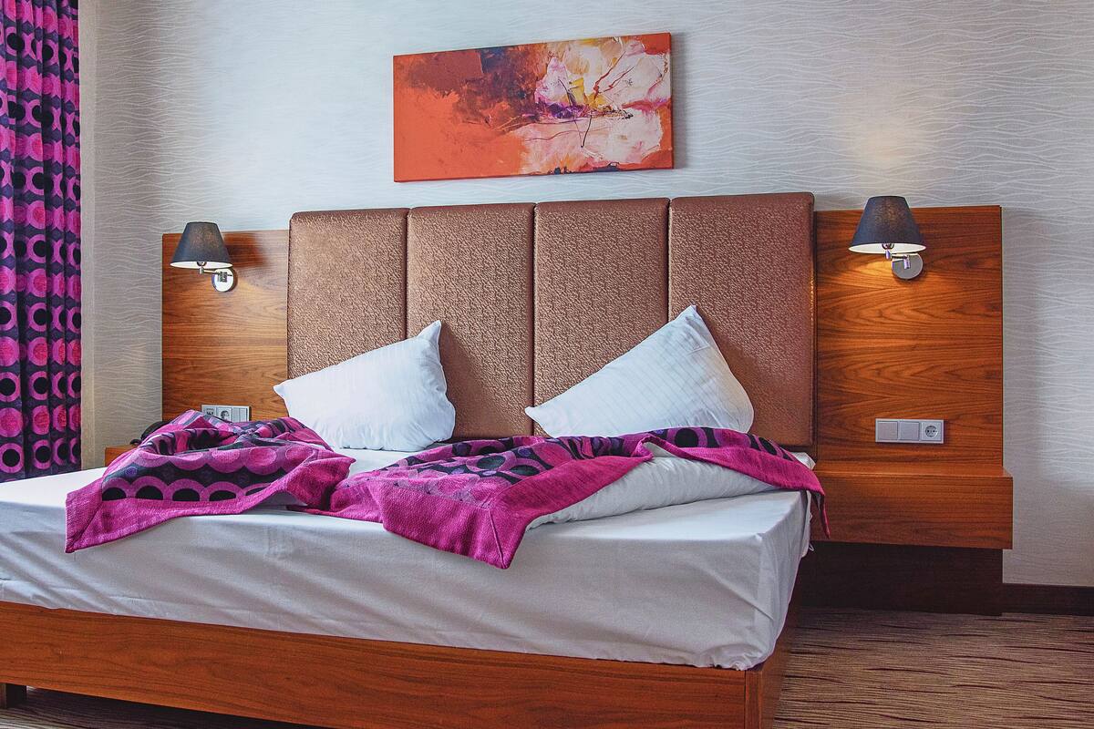 Quarto de hotel com cabeceira de madeira, duas luminárias de cabeceira, cama com roupa de cama branca e um cobertor colorido.