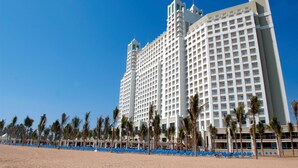 Um hotel grande com vários andares, localização à beira-mar e uma fileira de palmeiras ao primeiro plano.