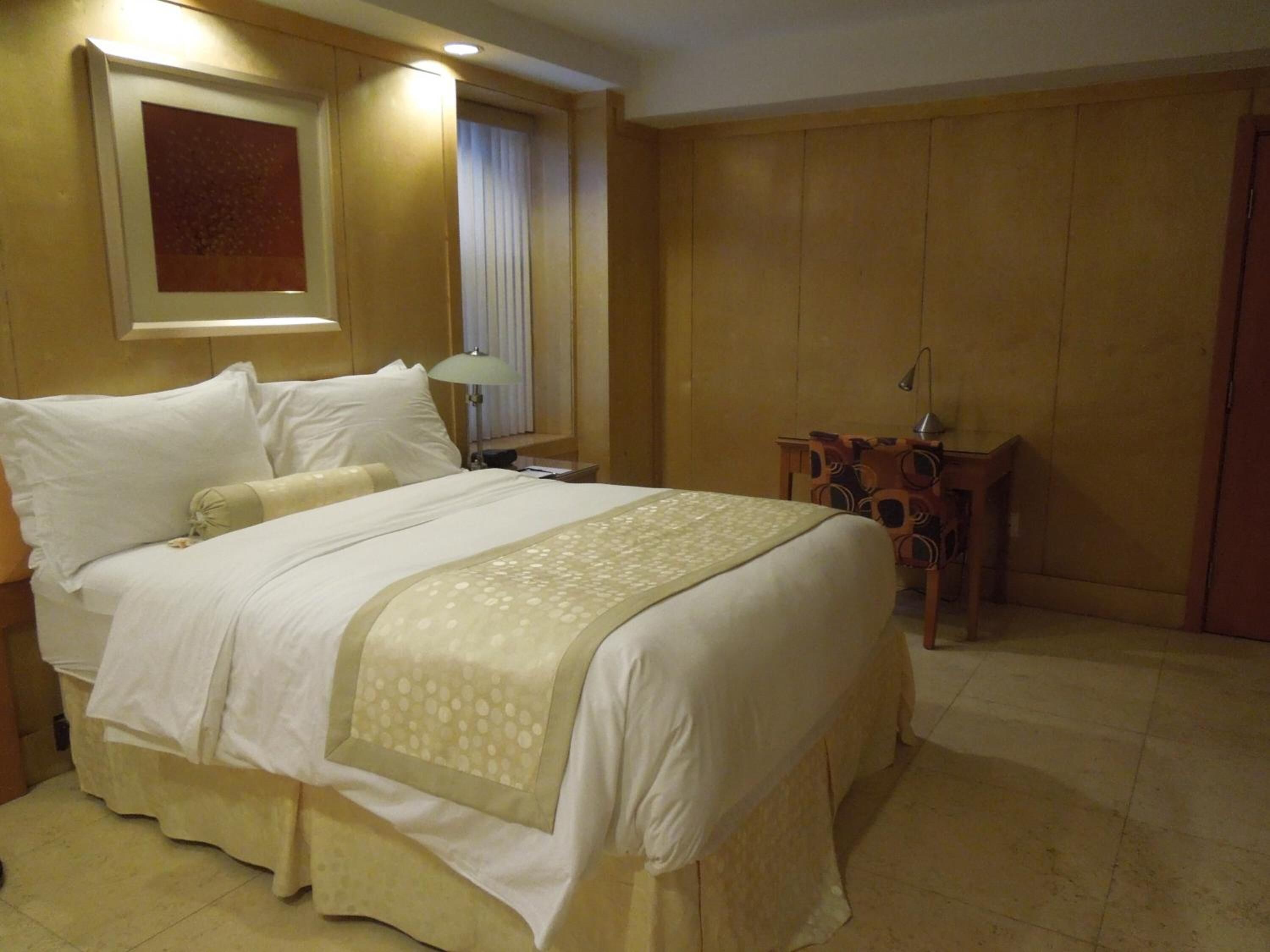 Cama bem arrumada com colcha estampada e almofadas, mesa de cabeceira com abajur e uma cadeira de madeira em quarto com lambris e um quadro na parede.