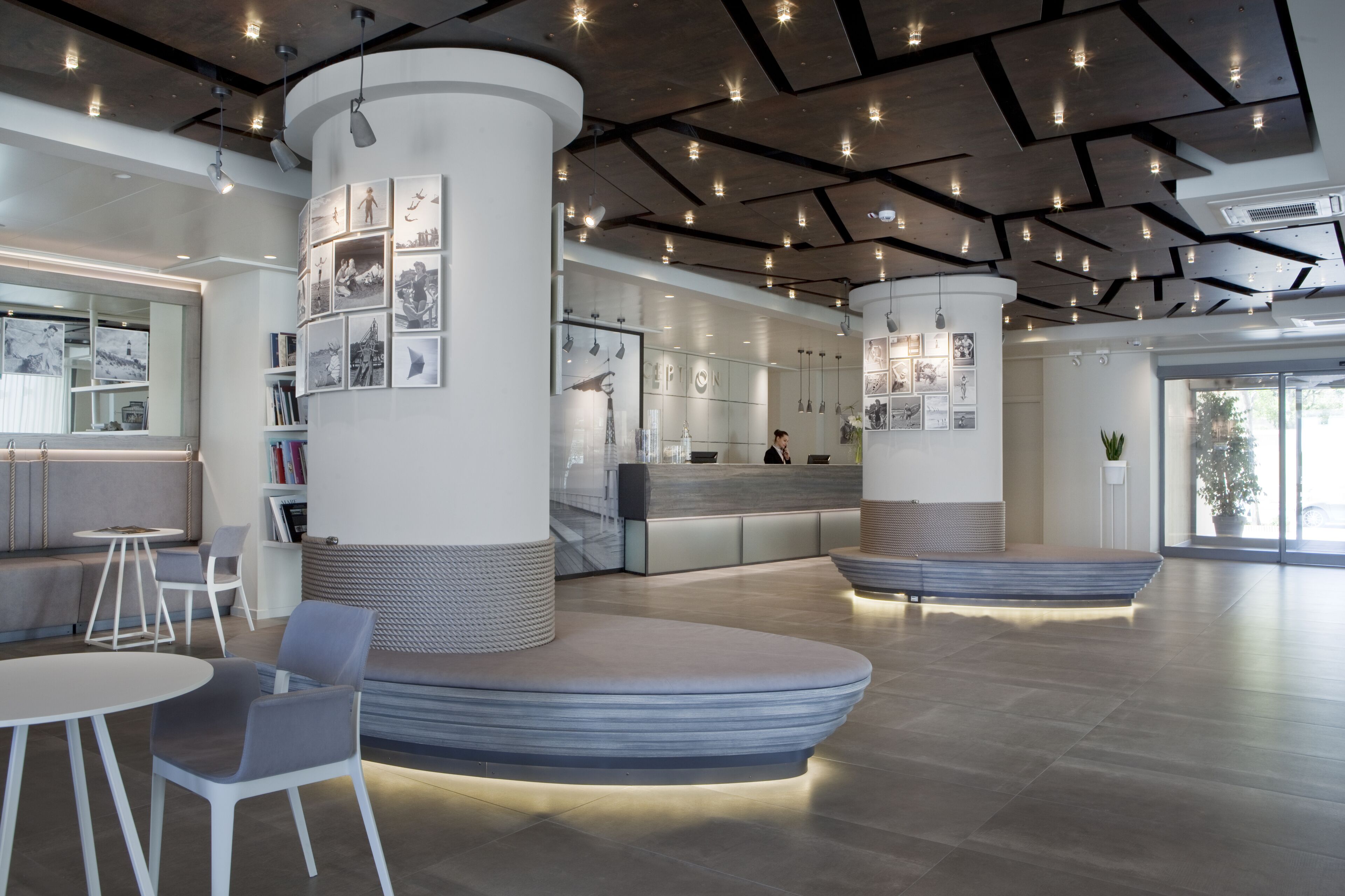Um lobby moderno com um balcão de recepção central, áreas de estar e um teto com vigas aparentes e iluminação embutida.