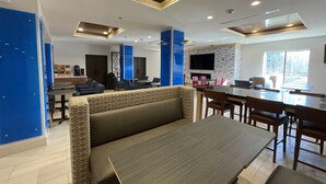 Uma cozinha moderna com TV integrada, sala de jantar com mesa grande e cadeiras, e um espaço de estar com sofá e um banco estampado.