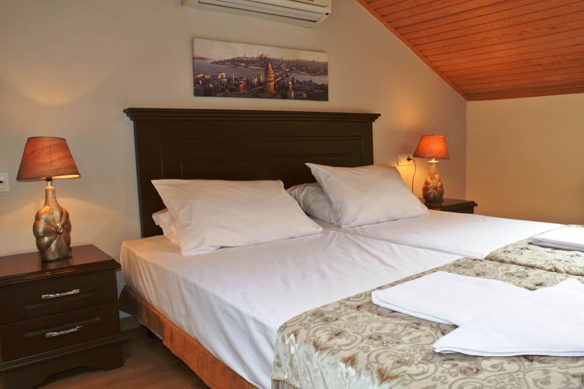 Cama bem arrumada com lençóis brancos, duas luminárias de cabeceira e uma pintura com vista para a cidade acima da cabeceira.