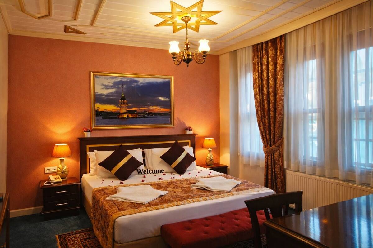 Quarto de hotel com cama, luminárias de cabeceira, luminária no teto em formato de estrela, pintura de um farol, mesa de jantar e uma cadeira vermelha.