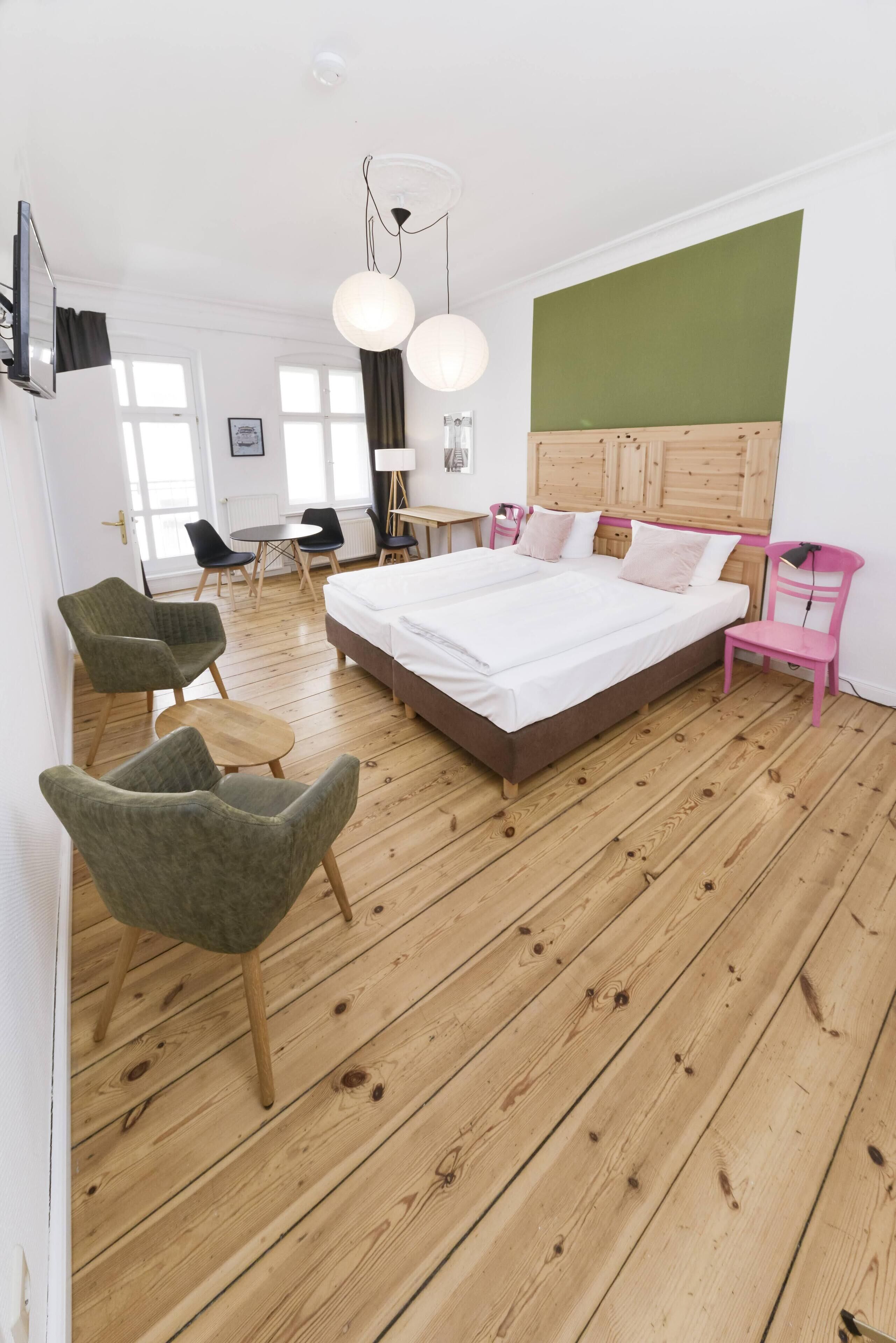 Quarto de hotel moderno com cama grande, piso de madeira, parede verde e área de refeições com cadeiras e mesa.