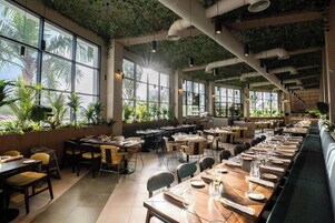 Um restaurante com grandes janelas, plantas dentro e várias mesas para refeições.