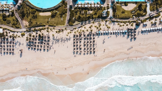 Vista aérea de uma praia com fileiras de palmeiras e cadeiras de praia, piscina e um edifício com telhado curvo.