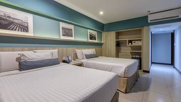 Quarto de hotel com duas camas, um guarda-roupa de madeira e uma pequena mesa de trabalho com um telefone e uma garrafa.