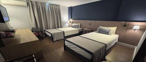 Quarto de hotel com duas camas de solteiro, uma escrivaninha com cadeira, televisão e janela com cortinas.