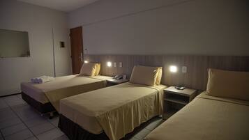 Quarto de hotel com três camas de solteiro, uma mesa de cabeceira com abajur e um espelho na parede.